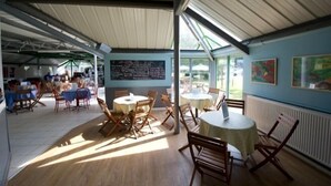 Um espaço de refeições interno com mesas e cadeiras, um menu em lousa e grandes janelas que permitem a entrada de luz natural.