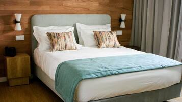 Quarto de hotel moderno com cama cinza, roupa de cama branca e manta azul-esverdeada. Há duas mesas de cabeceira com abajures e uma cabeceira de madeira.