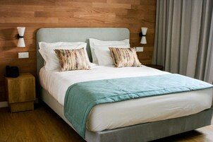 Quarto de hotel moderno com cama cinza, roupa de cama branca e manta azul-esverdeada. Há duas mesas de cabeceira com abajures e uma cabeceira de madeira.