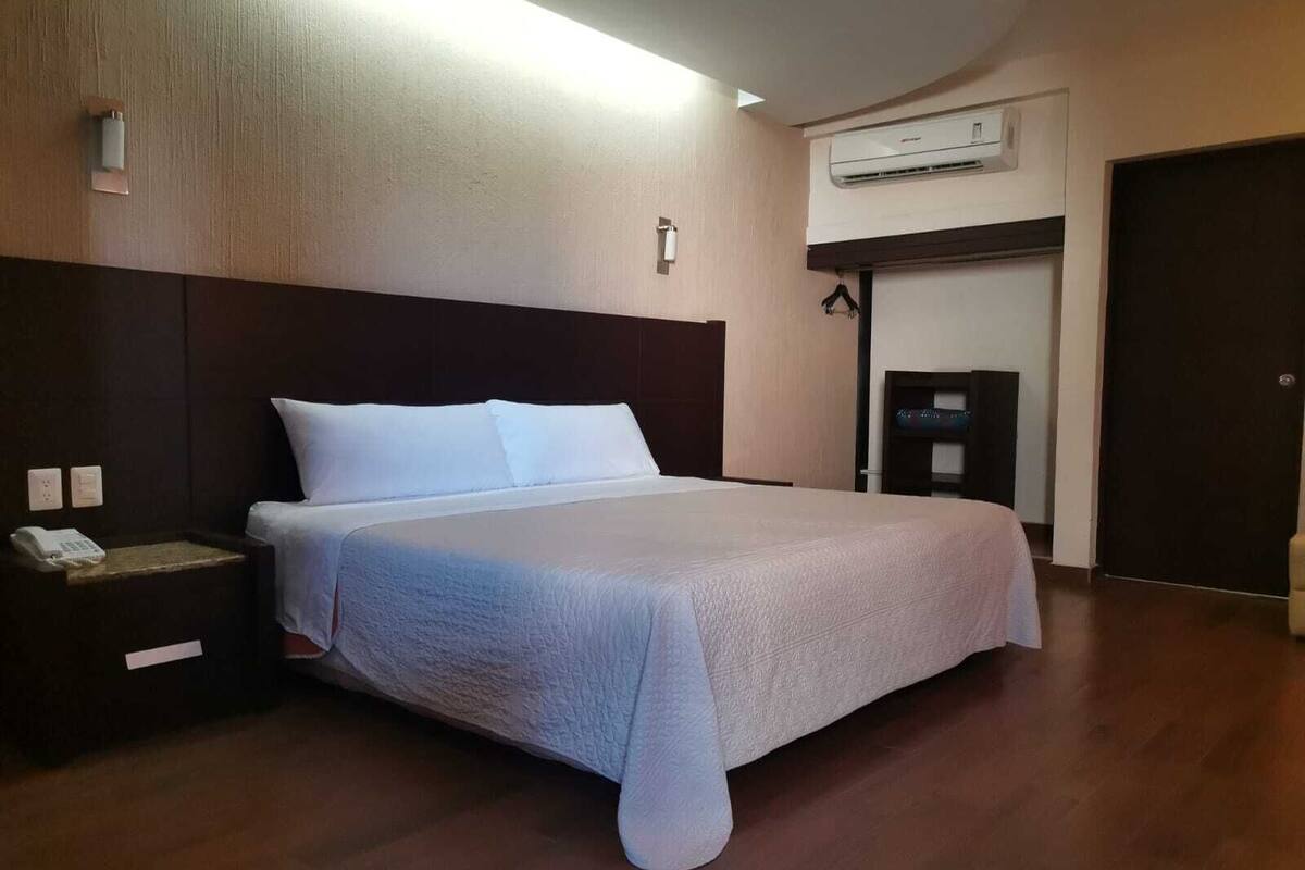Uma cama bem arrumada com edredom branco e travesseiros, uma mesa de cabeceira com um telefone e um aparelho de ar condicionado fixado na parede.