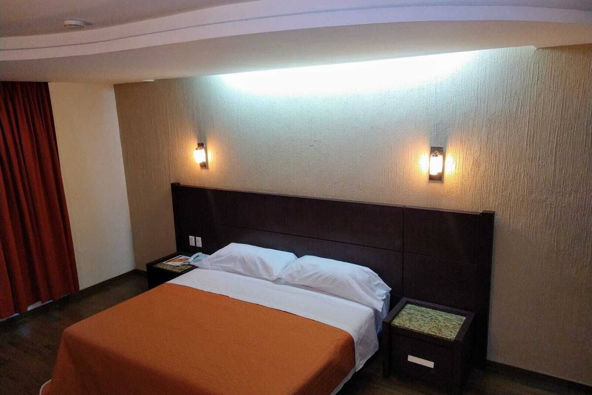 Quarto de hotel com uma cama grande, duas mesas de cabeceira e uma cabeceira com luzes fixadas na parede.