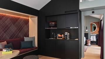 Quarto de hotel moderno com parede revestida de madeira escura, sofá vermelho com almofadas, mesa de madeira com um livro e uma cozinha com máquina de café e pia.