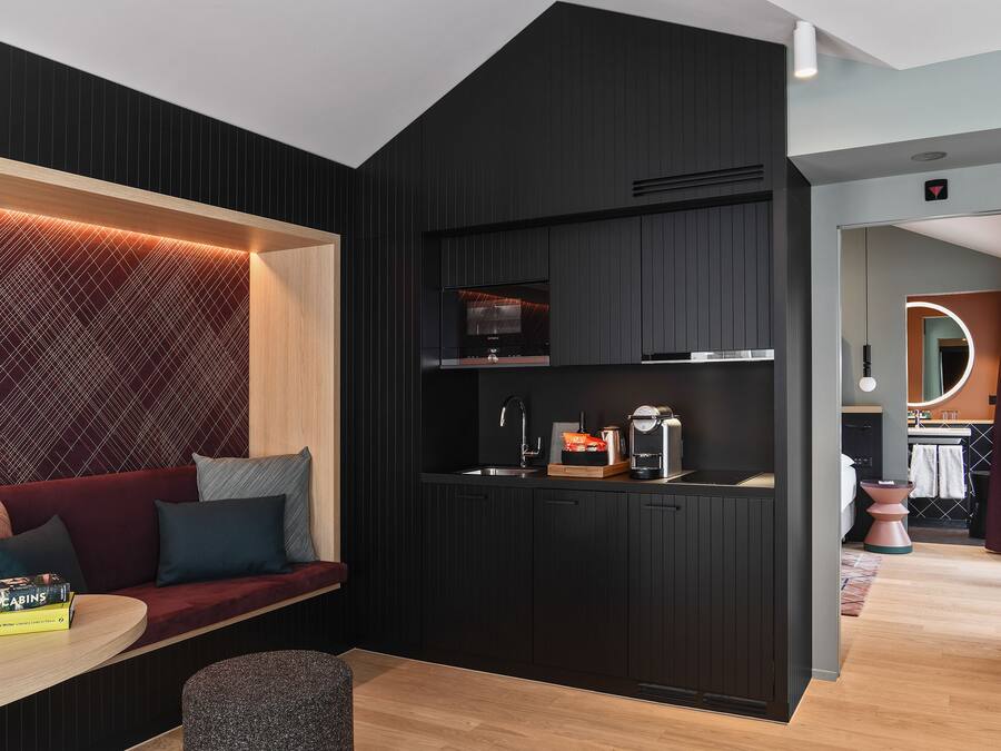 Quarto de hotel moderno com parede revestida de madeira escura, sofá vermelho com almofadas, mesa de madeira com um livro e uma cozinha com máquina de café e pia.