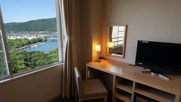 Quarto de hotel com escrivaninha, cadeira, TV e vista para um rio e paisagem urbana.