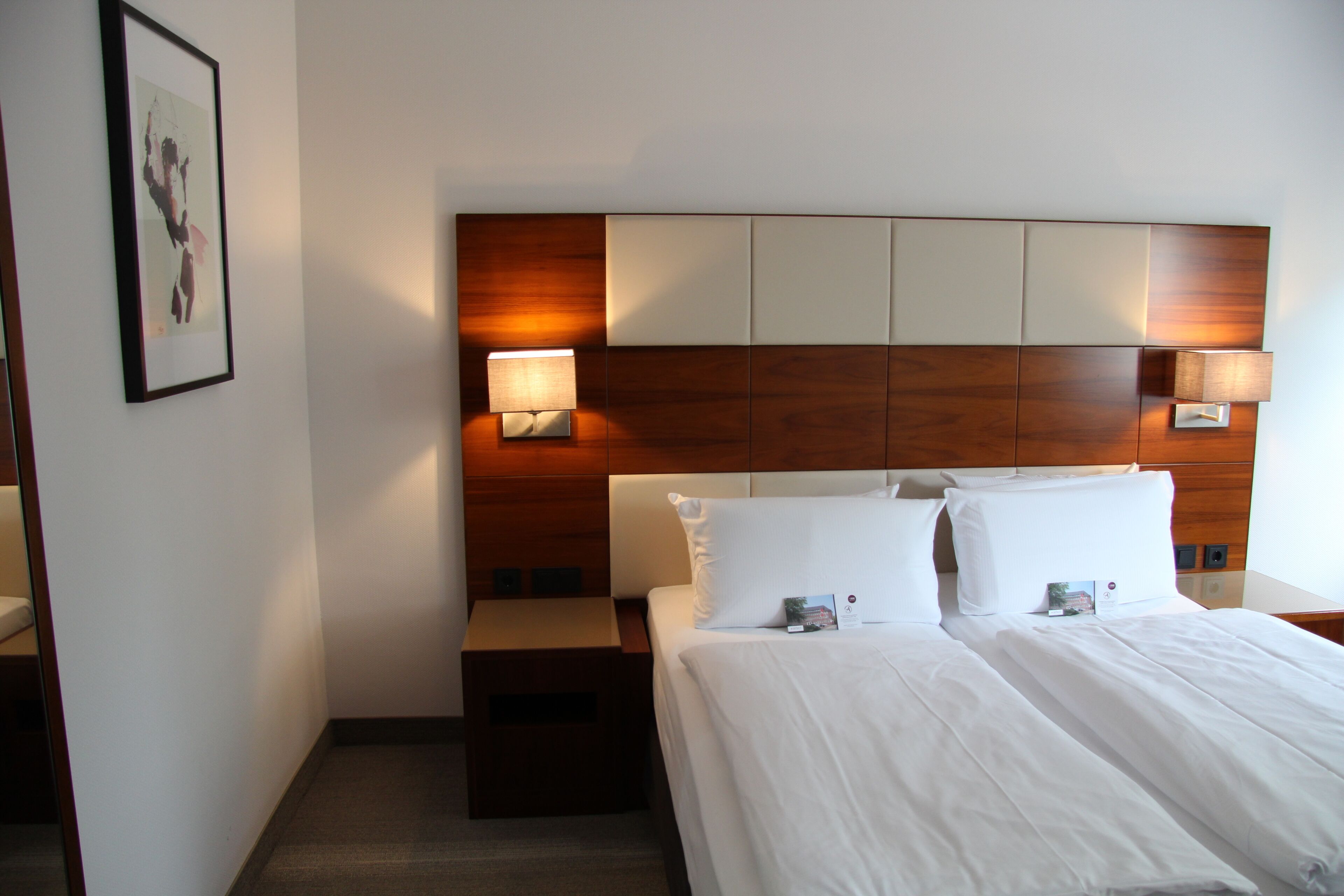 Quarto de hotel com cabeceira de madeira, duas mesinhas de cabeceira, cama com roupa de cama branca e duas luminárias fixadas na parede.