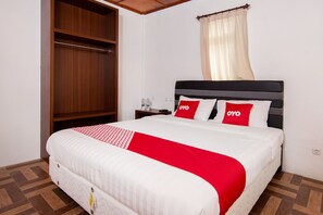 Cama bem arrumada com colcha vermelha e branca, duas almofadas com a marca 'OYO', um guarda-roupa de madeira e uma mesa de cabeceira com garrafas de água.