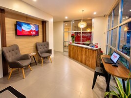 Uma área de recepção com balcão de madeira, duas poltronas cinzas, uma tela de TV e um laptop sobre uma mesa de madeira.