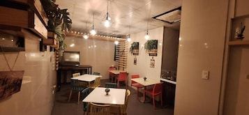 Uma sala de jantar com mesas e cadeiras, uma área de cozinha e uma estante com plantas.