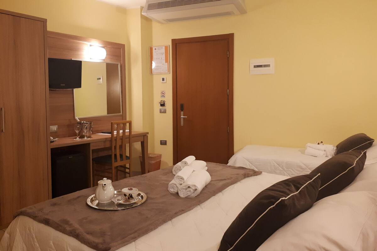 Quarto de hotel com uma cama, escrivaninha com cadeira, televisão e ar condicionado fixado na parede.