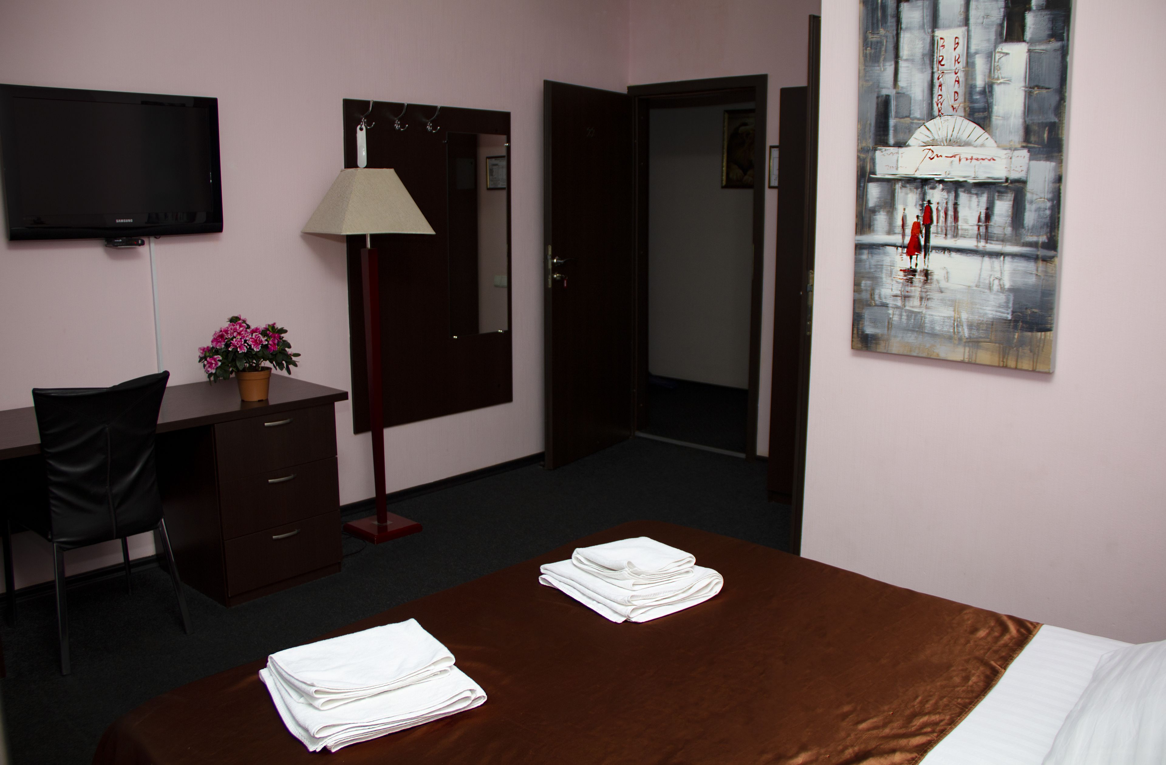 Quarto de hotel com cama, escrivaninha, cadeira, televisão, luminária e um quadro na parede.