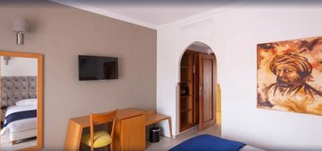 Quarto de hotel com cama, escrivaninha, cadeira, televisão, guarda-roupa e um quadro na parede.