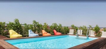 Piscina com deck de madeira, rodeada por bean bags coloridos e uma escada de metal.