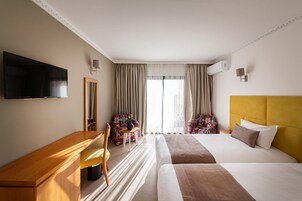 Quarto de hotel com cama, escrivaninha, cadeira, televisão e janela com cortinas.