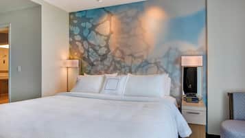 Quarto de hotel com uma cama grande, mesa de cabeceira com luminária, uma cadeira e parede com um mural texturizado.