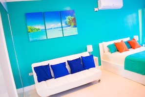 Quarto com sofá e cama brancos, paredes azul-turquesa e uma obra de arte com tema de praia.