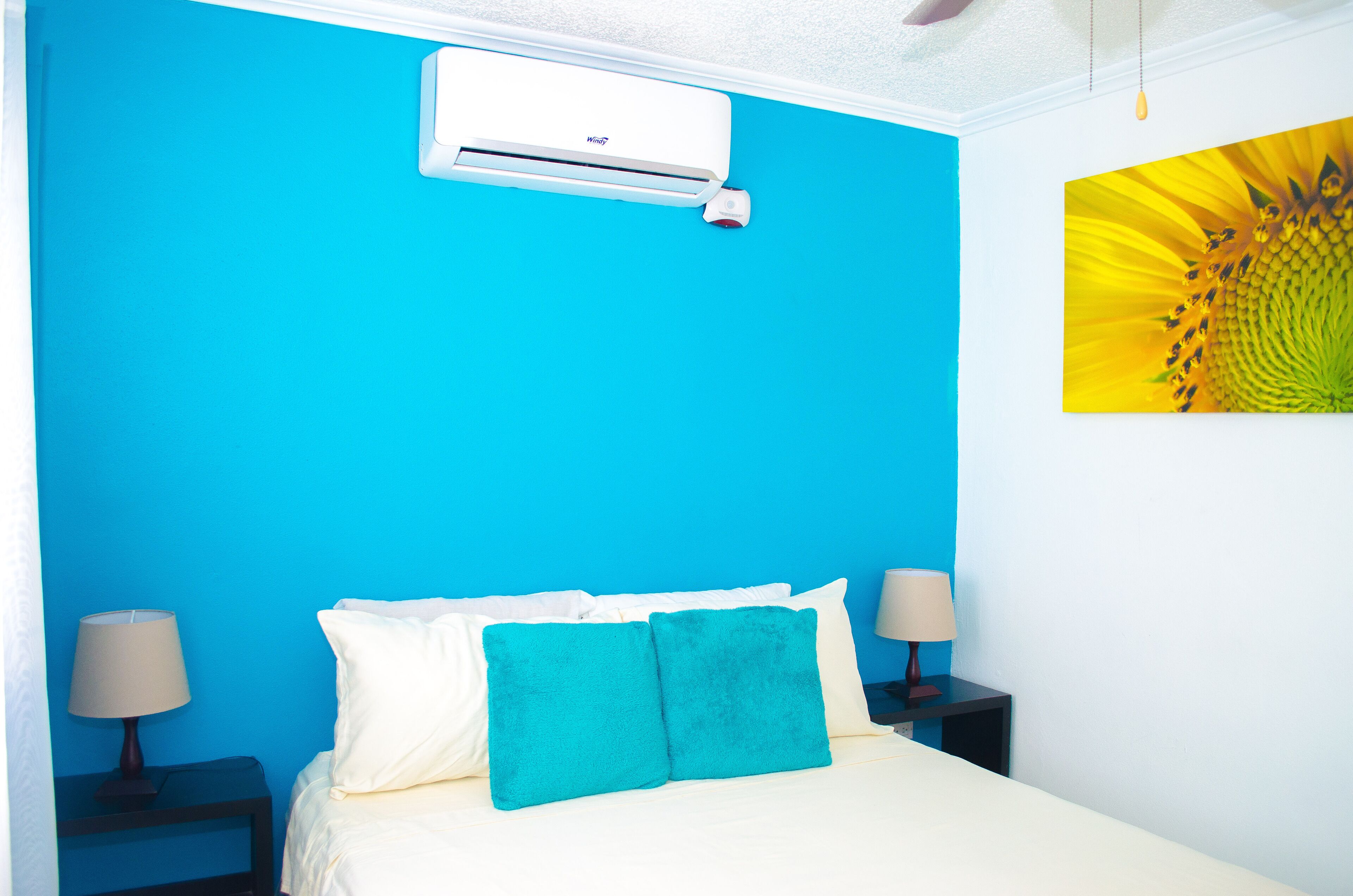 Quarto de hotel com parede azul vibrante, cama branca com duas almofadas brancas e duas almofadas decorativas azul-turquesa, unidade de ar condicionado branca e luminária fixa na parede a cada lado da cama.