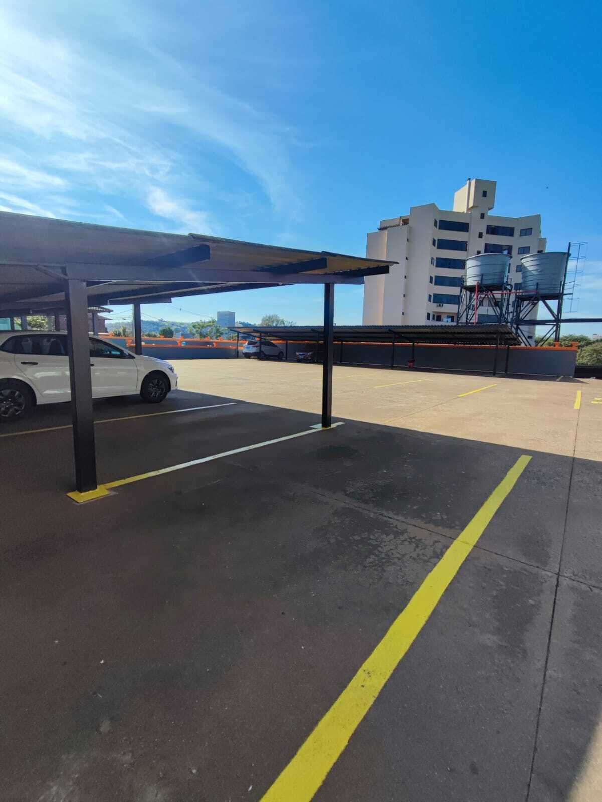Uma área de estacionamento com cobertura, um carro branco e um edifício de vários andares ao fundo.