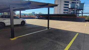 Uma área de estacionamento com cobertura, um carro branco e um edifício de vários andares ao fundo.