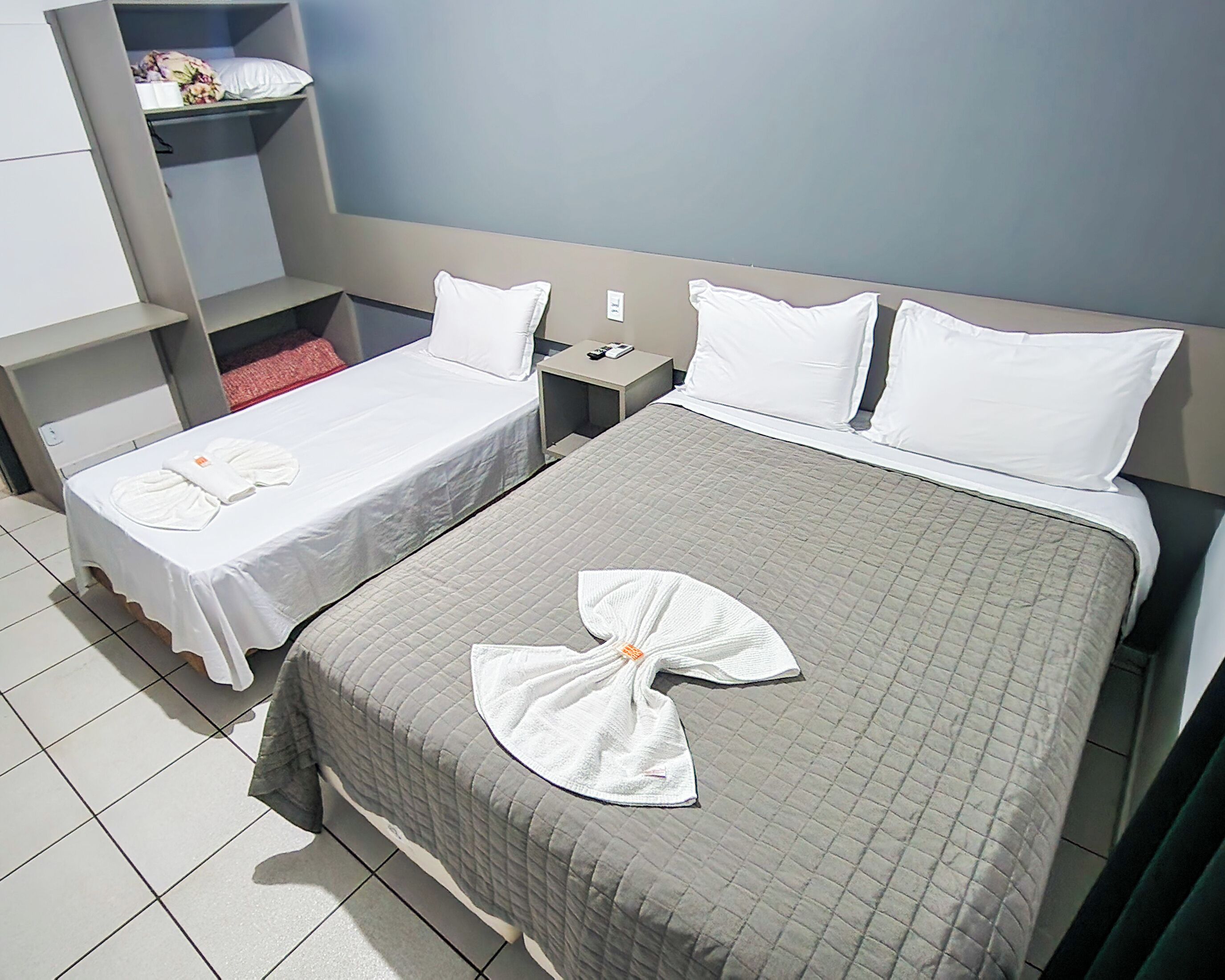 Quarto de hotel com duas camas, cada uma com roupa de cama e toalhas brancas. Há uma pequena mesa de cabeceira com um telefone e uma luminária. O quarto possui piso de azulejos e parede em tom claro.