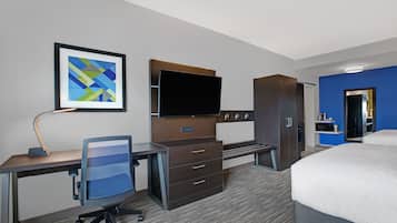 Quarto de hotel com escrivaninha, cadeira, televisão, guarda-roupa e cama.