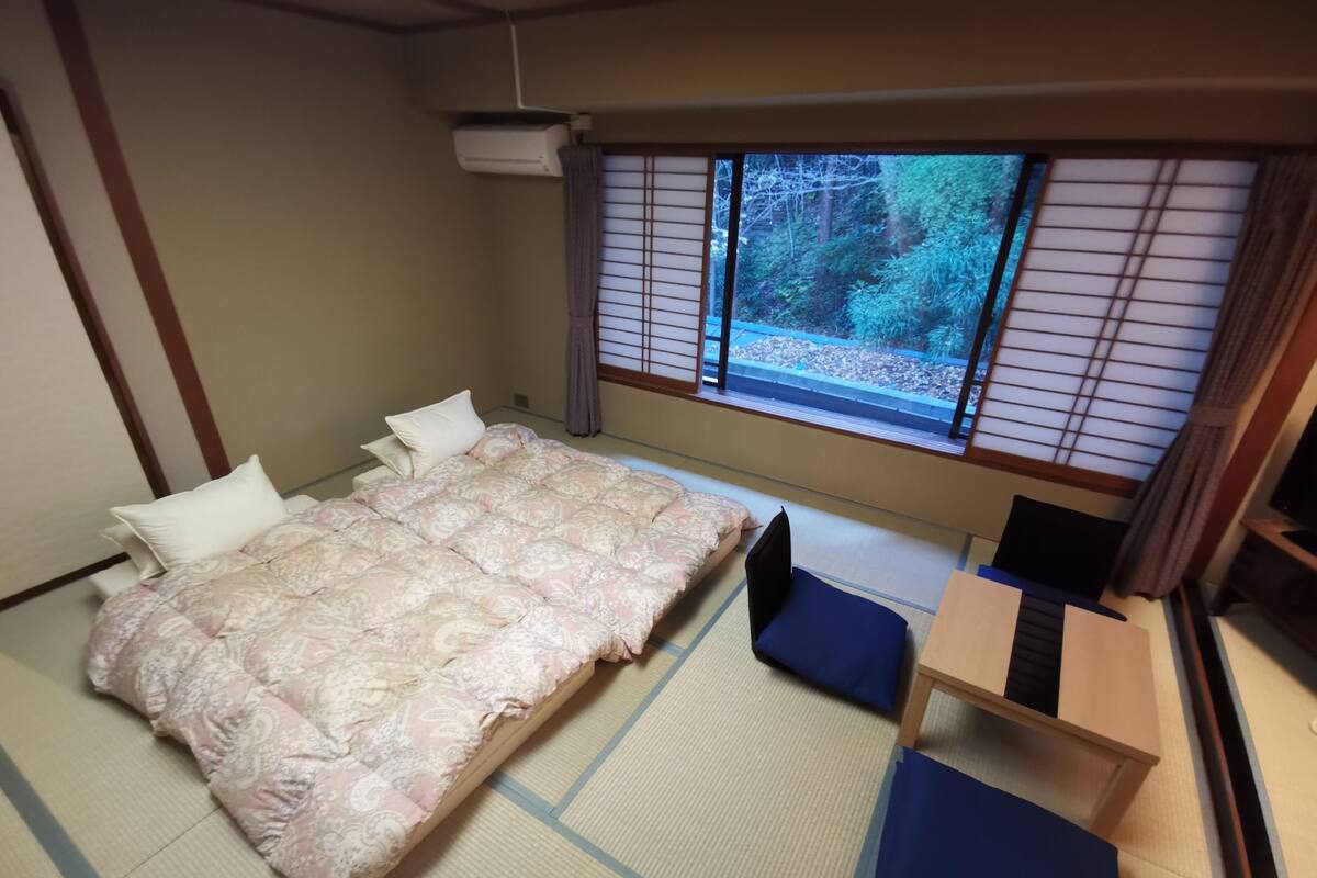 Quarto tradicional japonês com tatames, uma cama grande com colcha estampada, uma pequena mesa e vista para uma floresta através de biombos shoji.