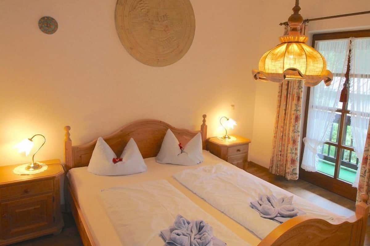 Uma cama de madeira com roupas de cama e toalhas brancas, duas mesas de cabeceira com abajures, um relógio de parede e uma janela com cortinas.