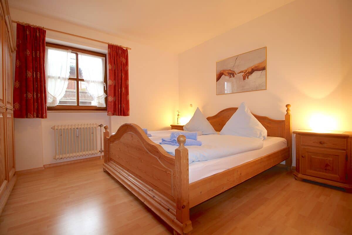 Quarto com cama de madeira, radiador, janela com cortinas vermelhas e mesa de cabeceira.
