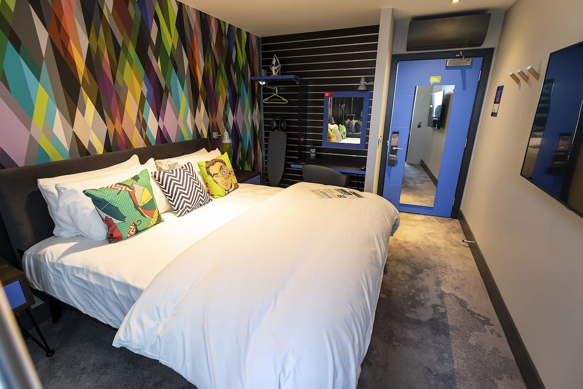 Quarto de hotel com cama grande, parede com padrão geométrico colorido, espelho e porta para o banheiro.