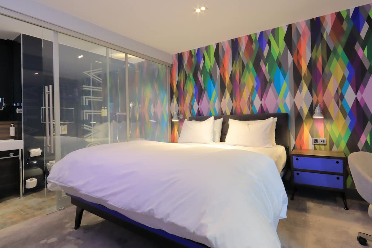 Quarto de hotel moderno com uma cama grande, box de vidro para o chuveiro, parede colorida com padrão geométrico, mesa de cabeceira com luminária e uma cadeira.