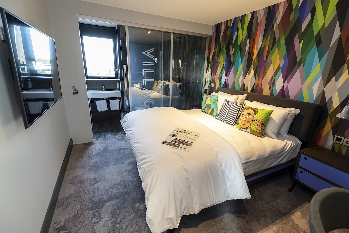 Quarto moderno com uma cama grande, porta de vidro que dá para uma varanda e um padrão geométrico colorido na parede.