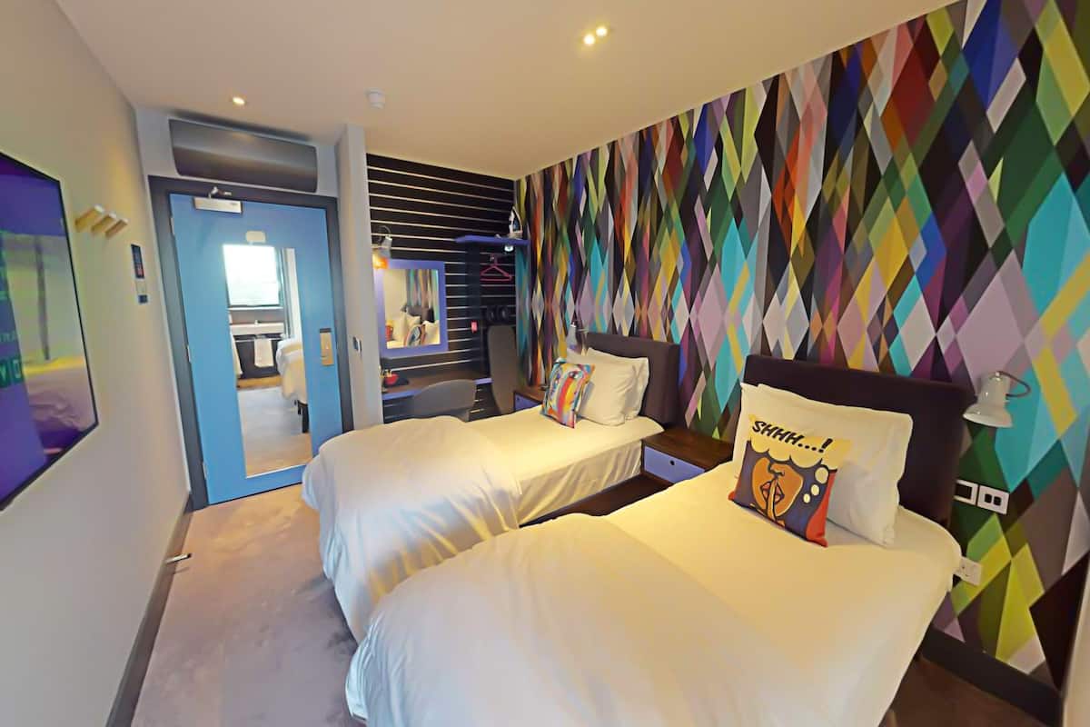 Quarto de hotel com duas camas, uma parede geométrica colorida, um espelho e uma pequena mesa de trabalho.
