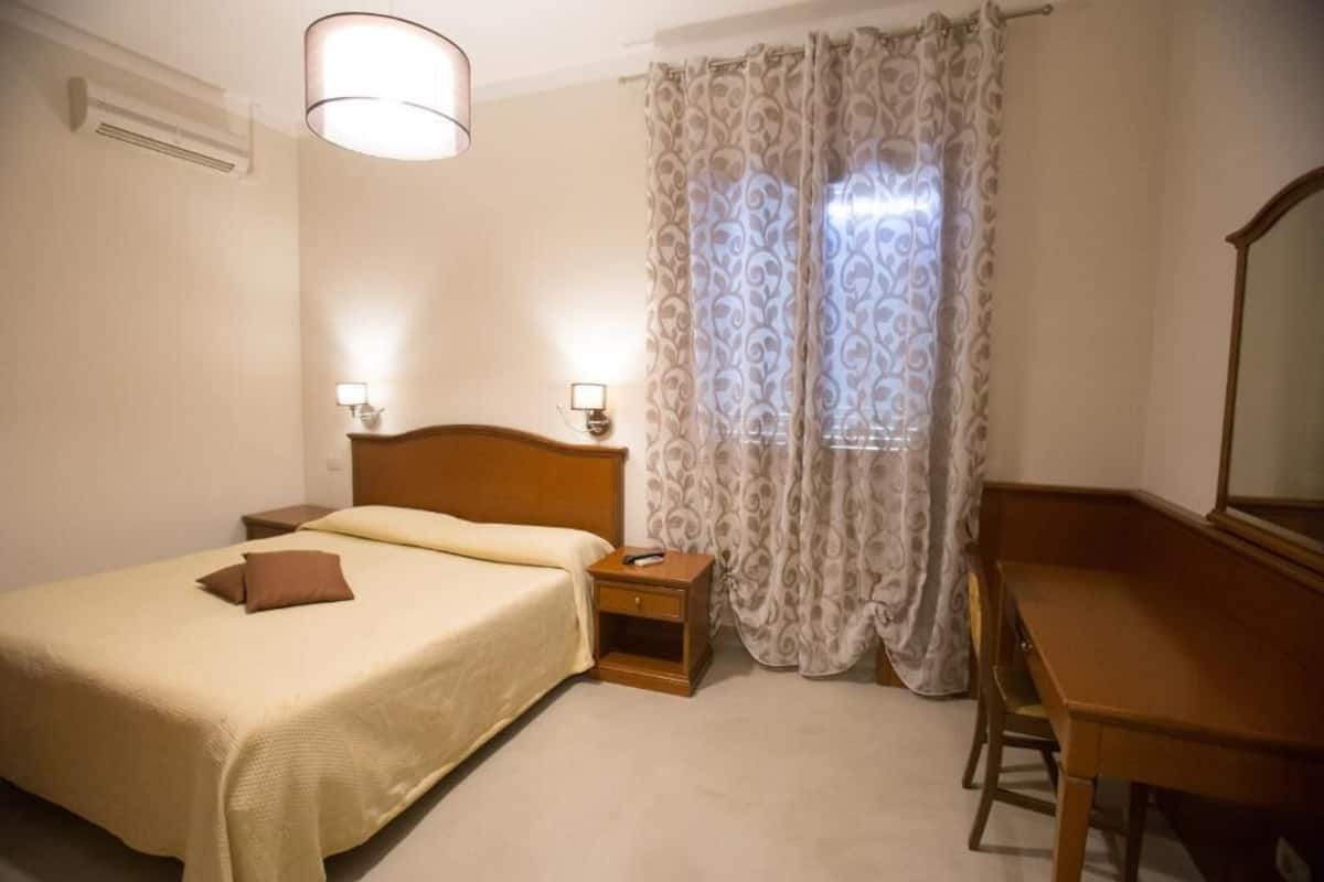 Quarto de hotel com cama, mesa de cabeceira, luminária, escrivaninha, cadeira e um espelho grande.