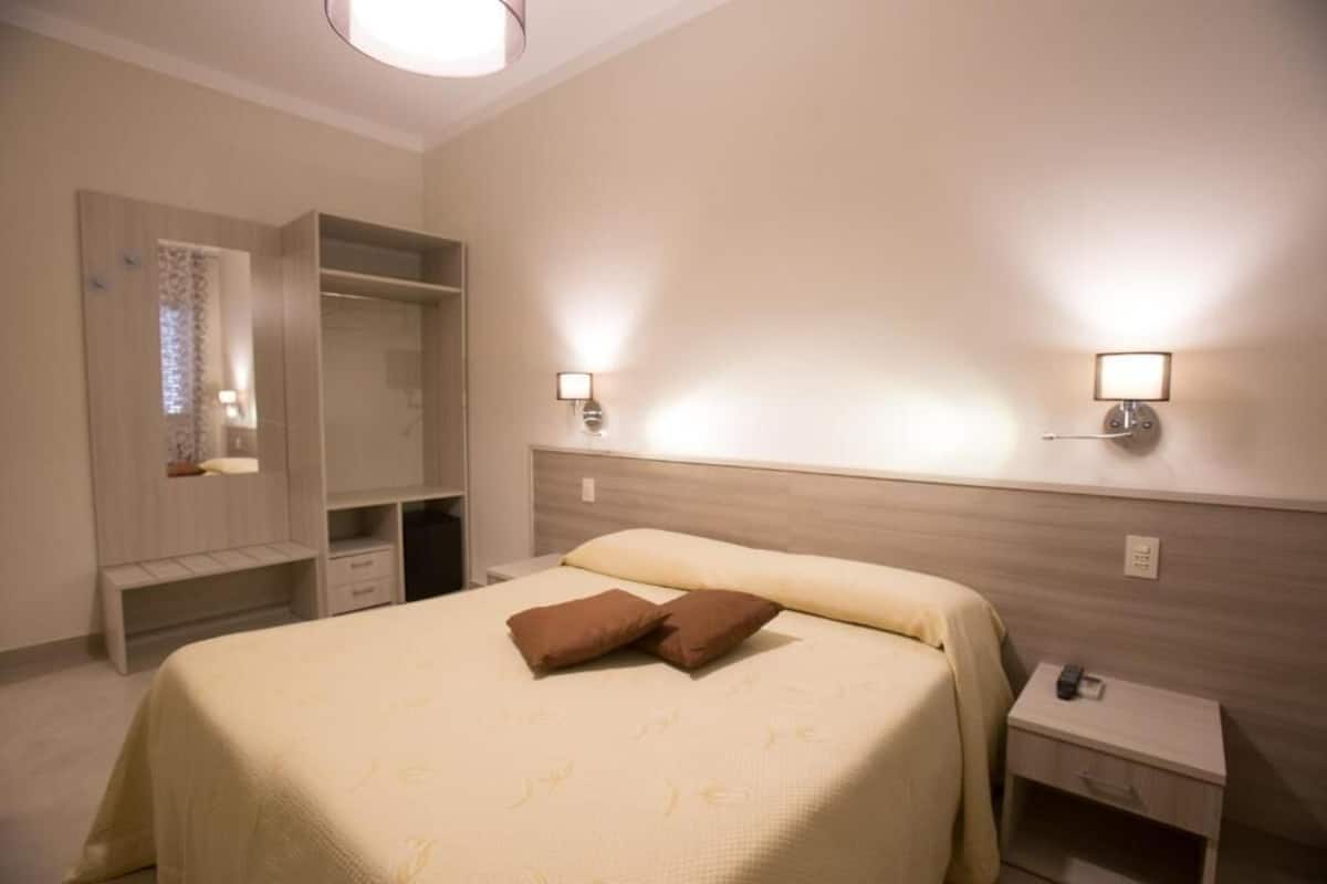 Quarto de hotel com cama, mesas de cabeceira, guarda-roupa e luminárias fixadas na parede.