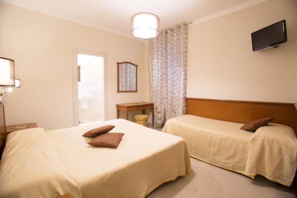 Quarto de hotel com duas camas de solteiro, televisão na parede, mesa de cabeceira com abajur e janela com cortinas.