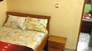 Quarto pequeno com uma cama de solteiro, cabeceira de madeira, mesa de cabeceira com gavetas e colcha estampada.