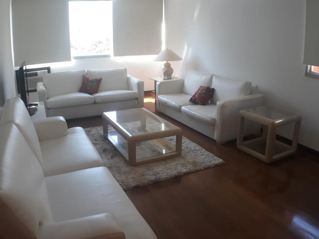 Uma sala de estar com um sofá branco, uma mesa de centro de vidro e uma pequena mesa lateral.