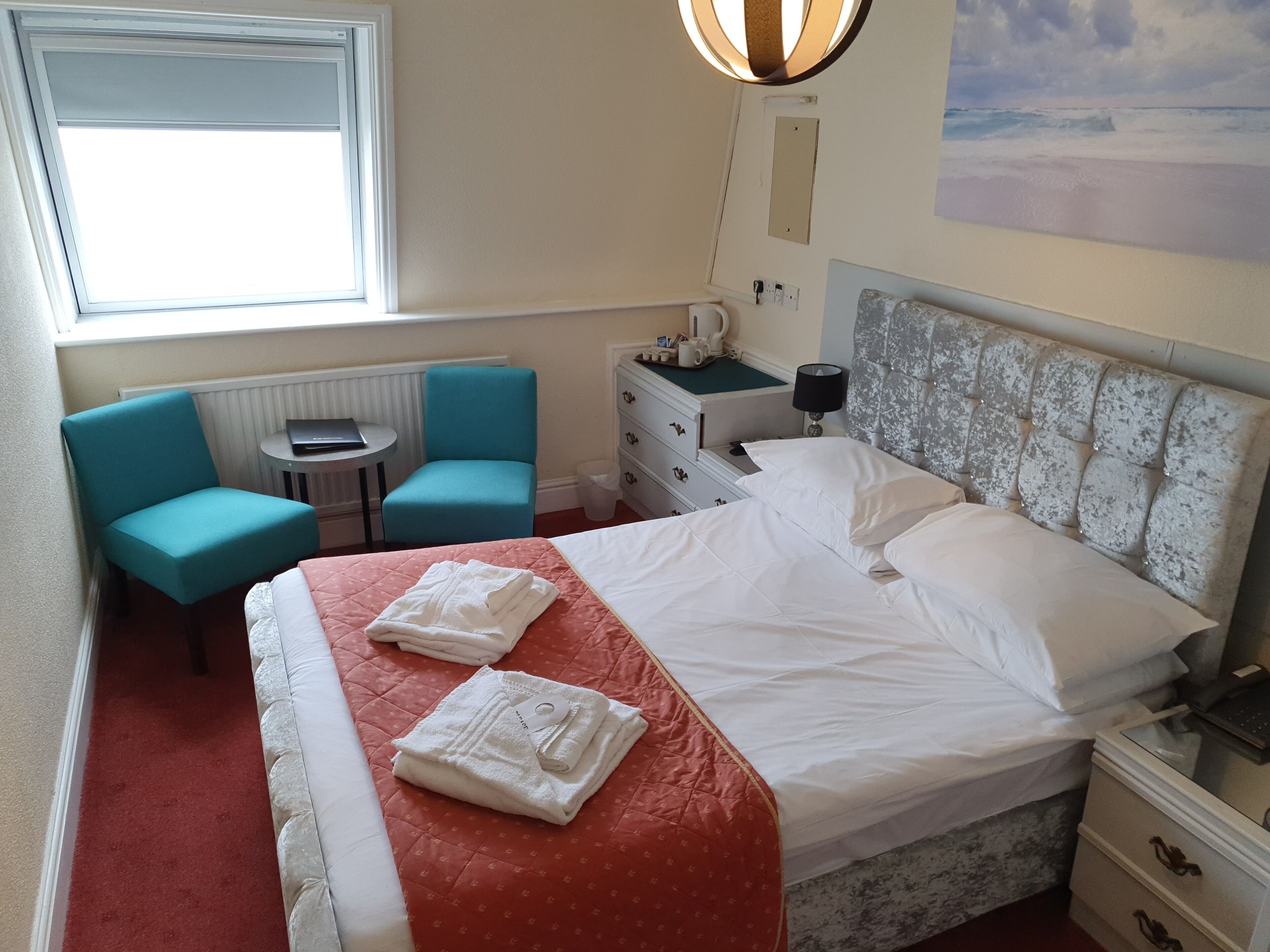 Quarto de hotel com uma cama, escrivaninha, cadeira, mesa de cabeceira e uma janela.