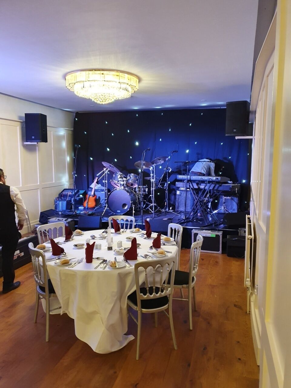 Uma sala de jantar com mesas redondas preparadas para uma refeição, um palco com instrumentos musicais e um candelabro.