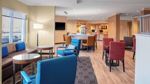 Um saguão de hotel com área de estar, com cadeiras azuis e vermelhas, uma pequena mesa redonda e um sofá com almofadas azuis. Há uma recepção ao fundo, com balcão e uma televisão presa à parede.