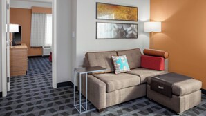 Quarto de hotel com sofá, apoio para os pés e uma pequena mesa lateral. Há uma televisão e um abajur sobre a mesa. O quarto possui um tapete estampado e uma porta que dá para outro cômodo.