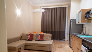 Um espaço de estar compacto com sofá, duas mesinhas, cozinha equipada com frigorífico e micro-ondas, e porta de correr para uma varanda.