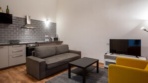 Uma sala de estar moderna com um sofá cinzento, uma mesa de centro, uma poltrona amarela, uma televisão de ecrã plano e uma área de cozinha com azulejos cinzentos e um micro-ondas.