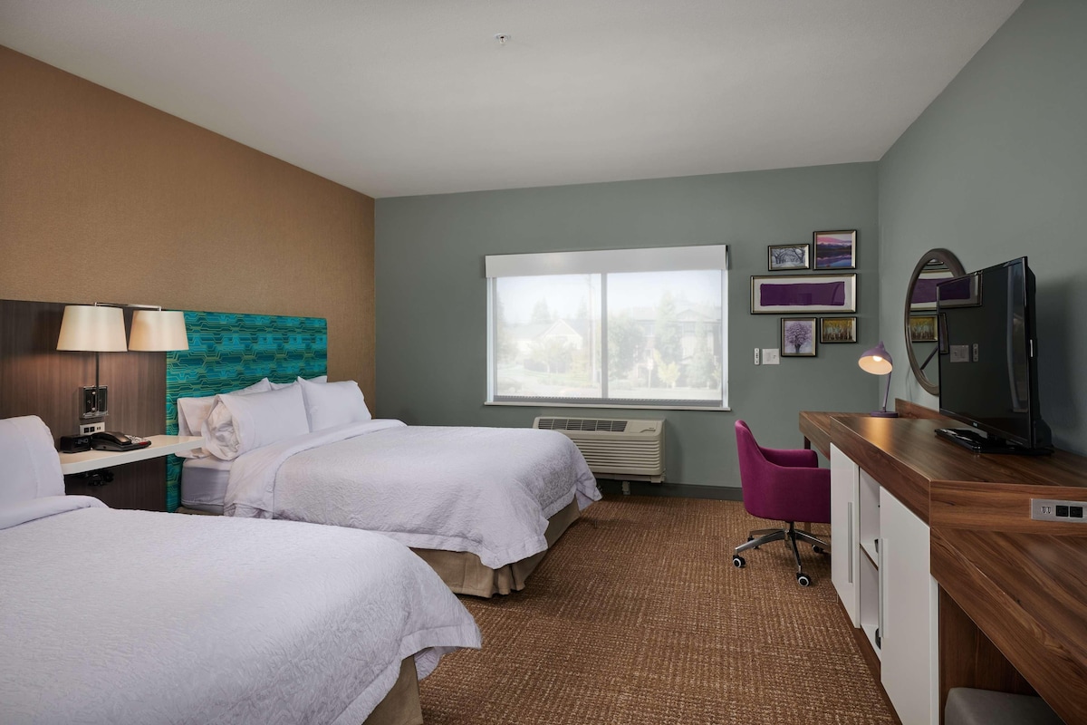 Quarto de hotel com duas camas, uma mesa, uma cadeira, televisão e janela.