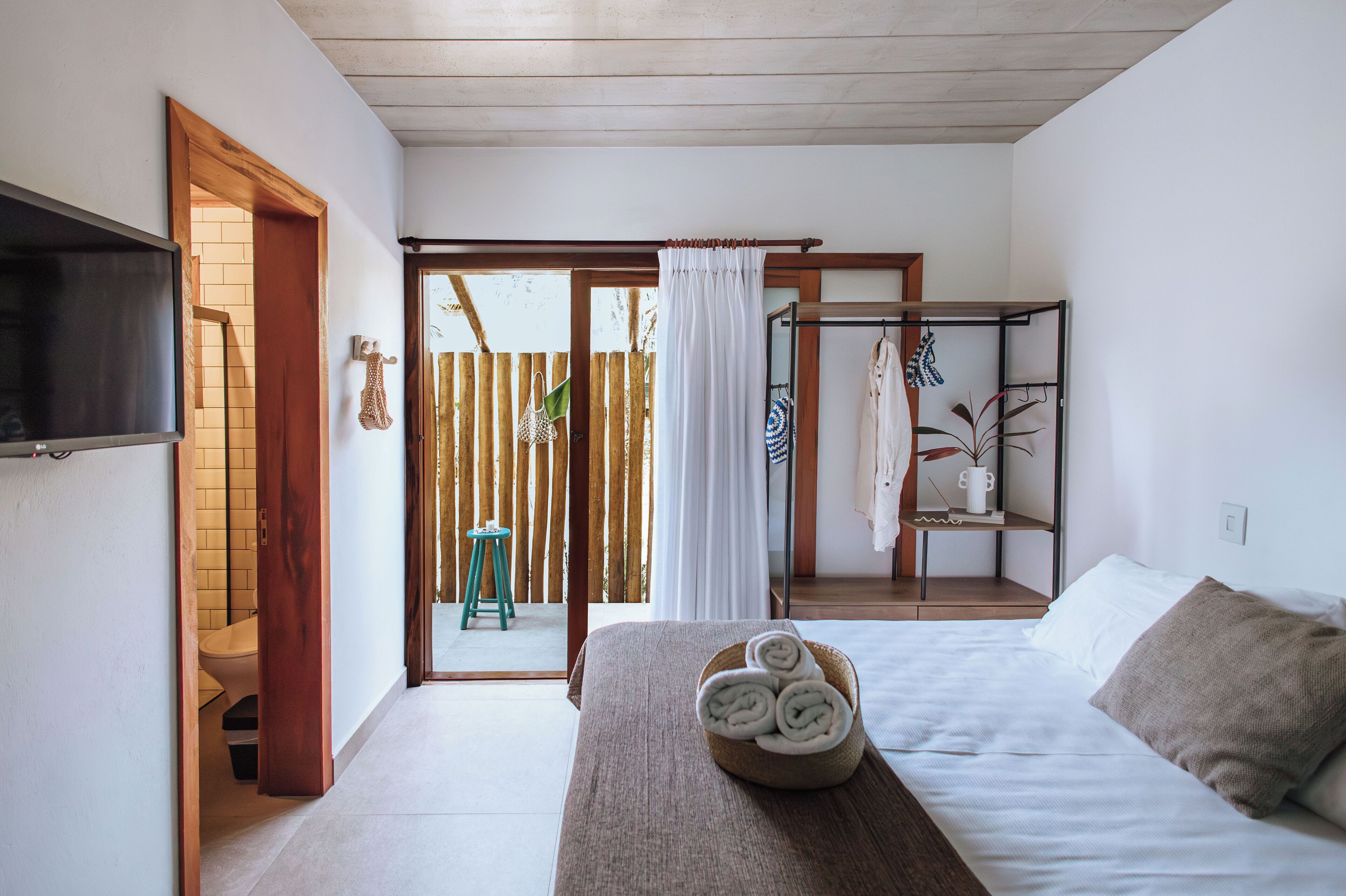 Quarto com cama, televisão, porta de madeira, um banquinho pequeno e vista para uma cerca de madeira lá fora.