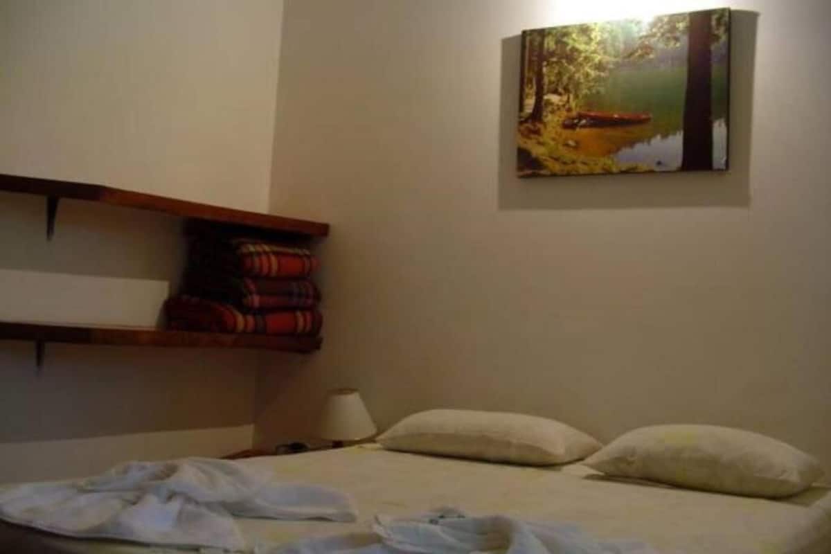 Quarto de hotel com cama, almofadas, um quadro na parede e uma estante com cobertores dobrados.