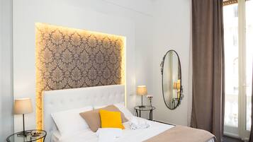 Quarto de hotel moderno com uma cama grande, cabeceira com padrão, espelho redondo e pequena mesa com abajur.