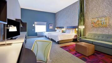 Quarto de hotel com cama, escrivaninha com computador, poltrona, sofá e tapete colorido.