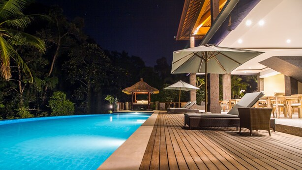 Piscina com espreguiçadeiras e guarda-sóis, rodeada por um deck de madeira e vegetação tropical.