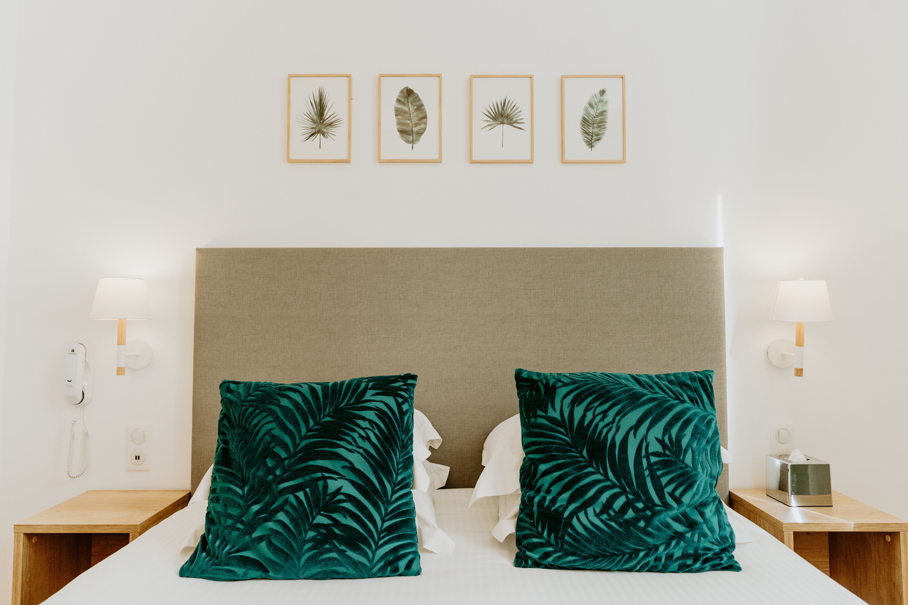 Cama bem arrumada com duas almofadas azul-turquesa, cabeceira bege e quatro folhas emolduradas na parede.