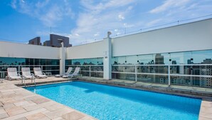 Piscina no terraço com água azul cristalina, rodeada por espreguiçadeiras e guarda-corpo de vidro.